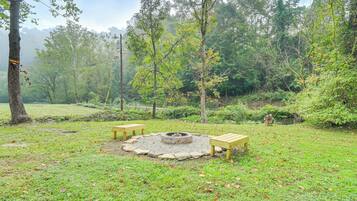 Uma fogueira circular com bancos, cercada por uma borda de pedra, situada em uma área gramada com árvores ao fundo.