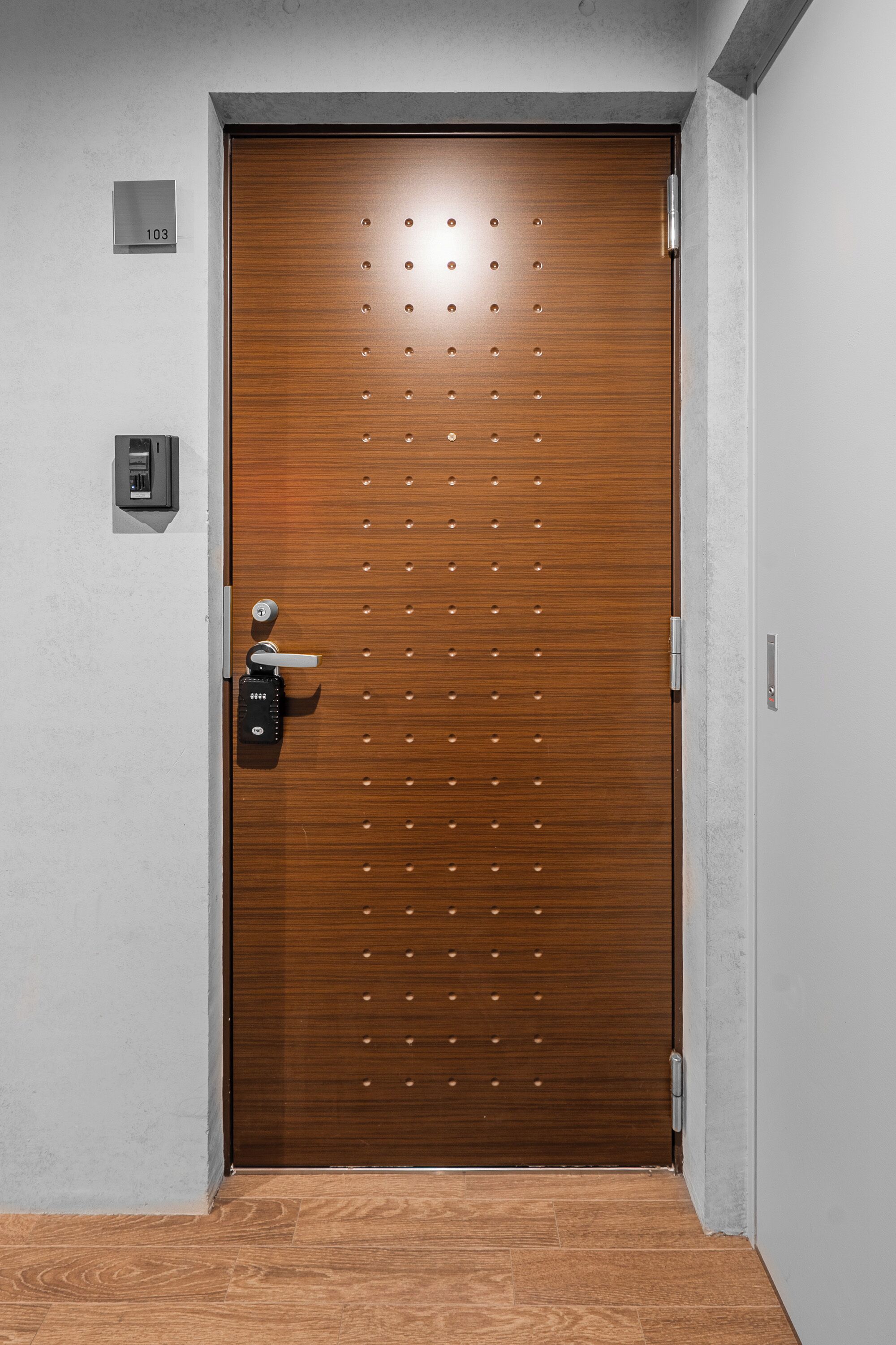 Uma porta de madeira com fechadura por teclado, uma pequena placa acima dela e um interruptor de luz na parede.