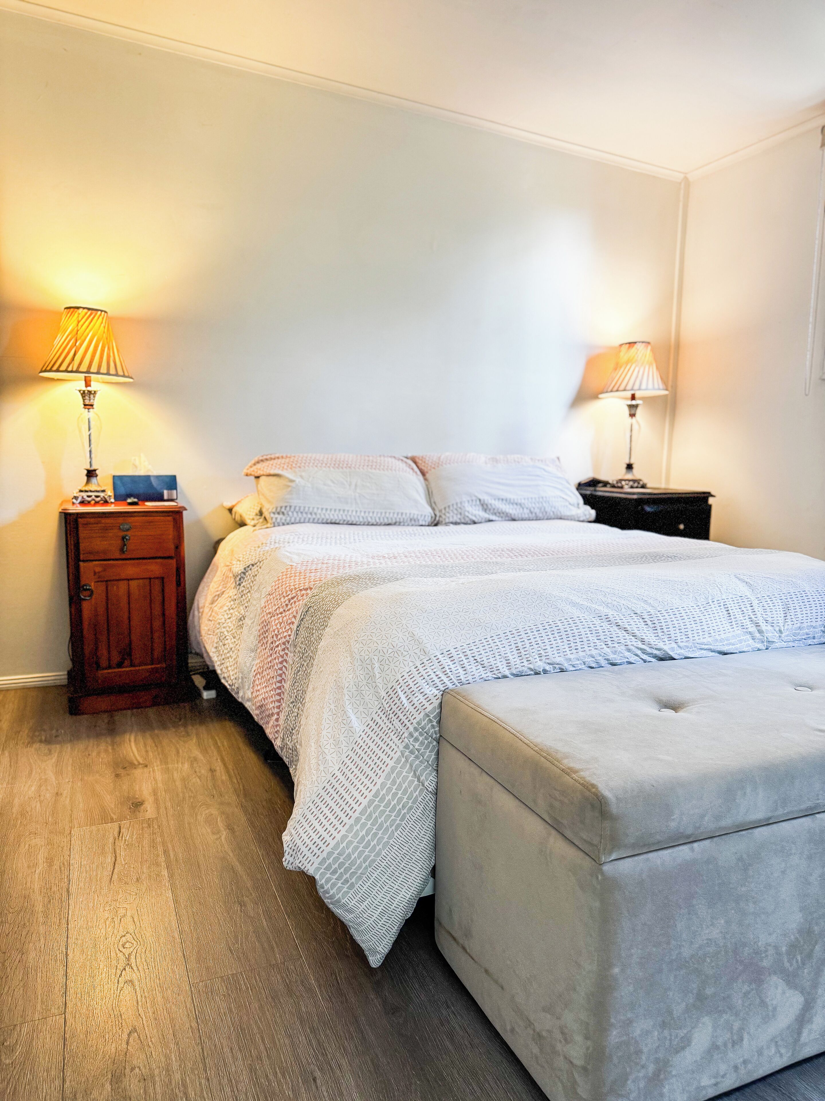 Um quarto com uma cama, duas mesas de cabeceira, um criado-mudo, piso de madeira e uma luminária fixada na parede.