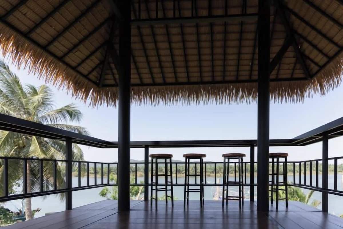 Um pavilhão aberto com telhado de palha com vista para um corpo de água, apresentando um espaço de bar com banquetas.