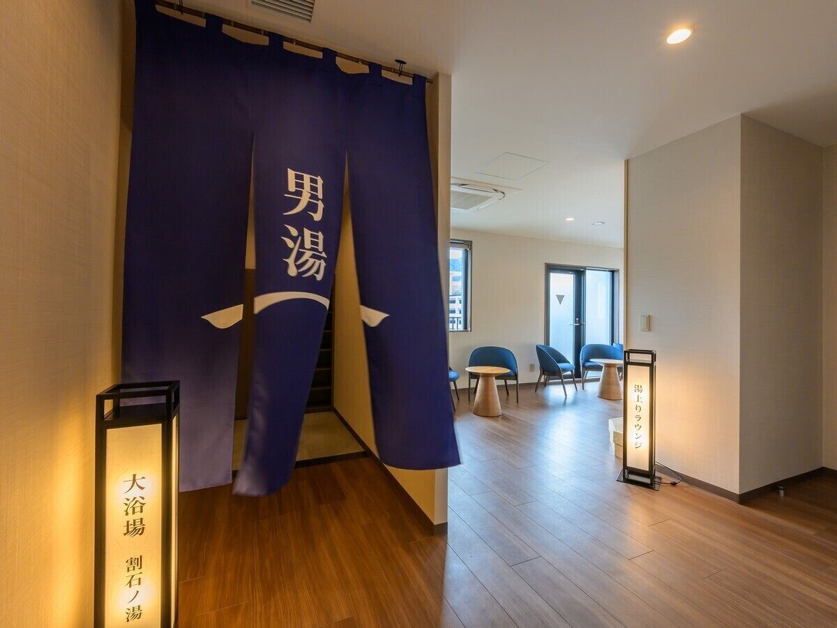 Corredor de hotel com piso de madeira, cortina azul com caracteres japoneses e um tradicional lampião japonês.