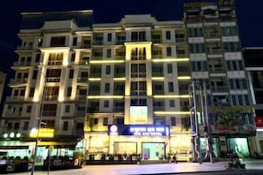 Hotel de vários andares com letreiros iluminados, entrada visível e rua em frente.