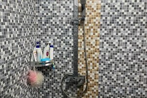 Área de duche com azulejos, um chuveiro, uma escova para o corpo pendurada e produtos de higiene numa prateleira.