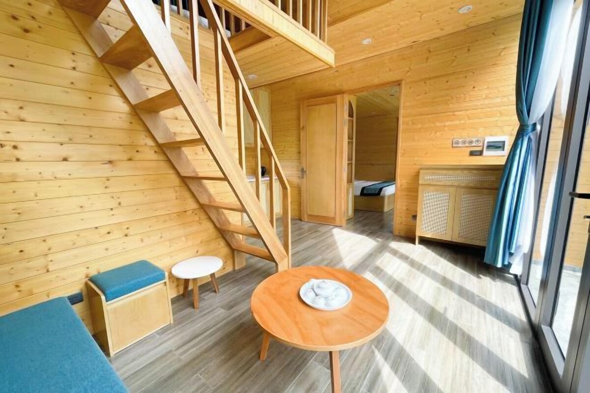 Interior em madeira com escada, mesa redonda com um prato e uma cama ao fundo.