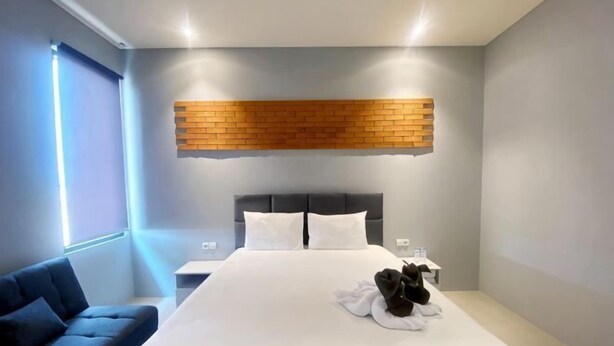 Quarto de hotel moderno com uma cama grande, um sofá azul, uma parede de tijolos e uma mesa de cabeceira com luminária.
