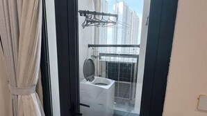 Uma pequena varanda com uma máquina de lavar branca, um varal de metal e vista para edifícios altos.