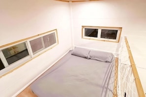 Quarto pequeno com cama de solteiro, janela, escada de madeira e parede branca.
