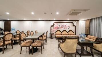 Um espaço de refeições de hotel com várias mesas e cadeiras, uma parede decorativa e uma área de estar com sofás e um vaso.