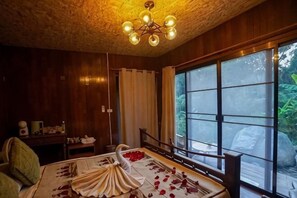 Quarto com cama, um candelabro, janela com cortinas e vista para o verde lá fora.