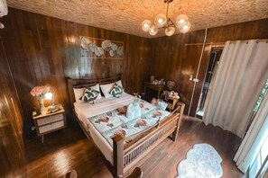 Quarto com piso de madeira, cama, mesas de cabeceira, abajur e um objeto decorativo na parede.