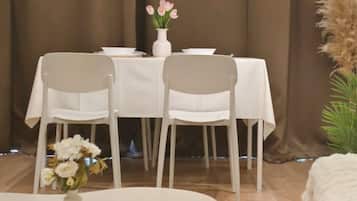 Uma área de refeições com uma mesa posta para quatro pessoas, cadeiras brancas e um vaso com flores.