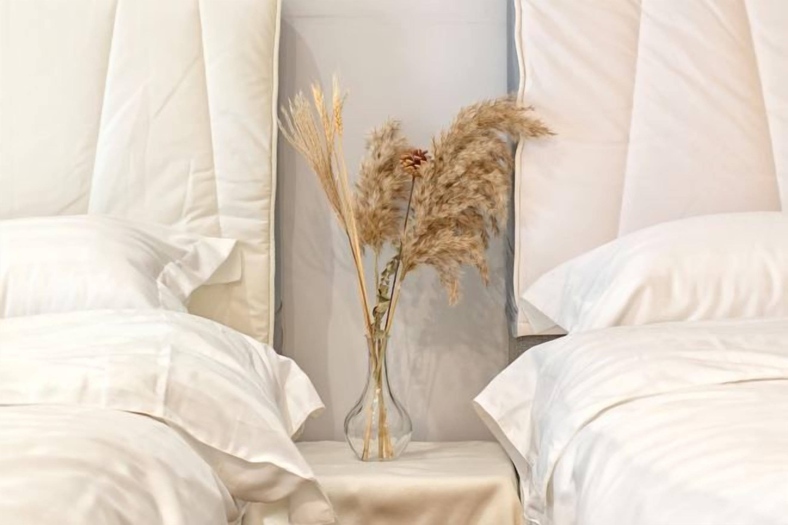 Uma cama com roupa branca, um vaso com plantas secas e uma parede em tom claro.