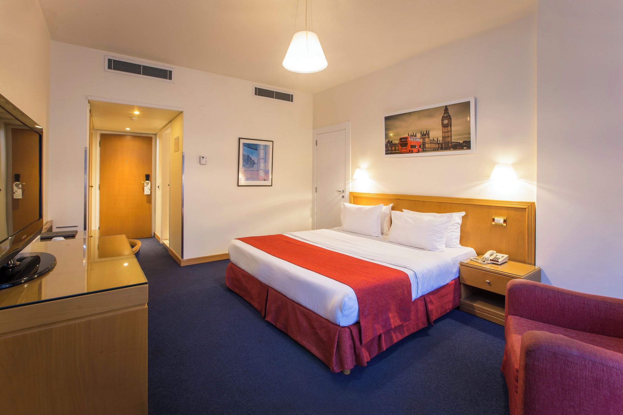 Quarto de hotel com cama, mesa de cabeceira, luminária, cadeira, escrivaninha e uma pintura de um horizonte urbano.