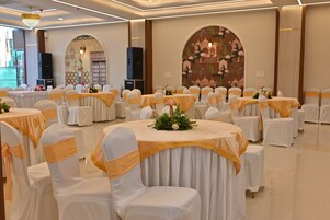 Uma sala de banquetes com mesas redondas, cadeiras brancas com fitas laranja e centros de mesa florais.