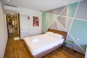 Quarto de hotel com piso de madeira, cama com roupa de cama branca, parede verde com padrões geométricos e um pequeno banquinho.