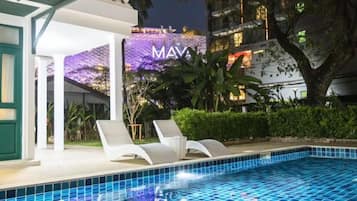Piscina com espreguiçadeiras, área de estar coberta e um edifício com letreiros iluminados ao fundo.