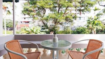 Uma varanda com uma mesa redonda e duas cadeiras de vime, com vista para um jardim com árvores e um edifício ao fundo.