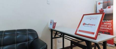 Quarto com sofá de couro preto, uma mesa com um certificado emoldurado e um cartaz promocional da RedDoorz.