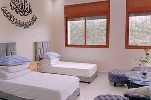 Quarto de hotel com duas camas, uma mesa pequena, um banquinho redondo e uma parede com caligrafia árabe.