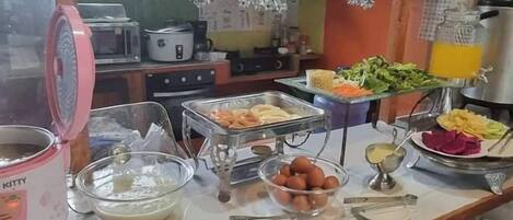Uma mesa de buffet com diversos alimentos e eletrodomésticos ao fundo.