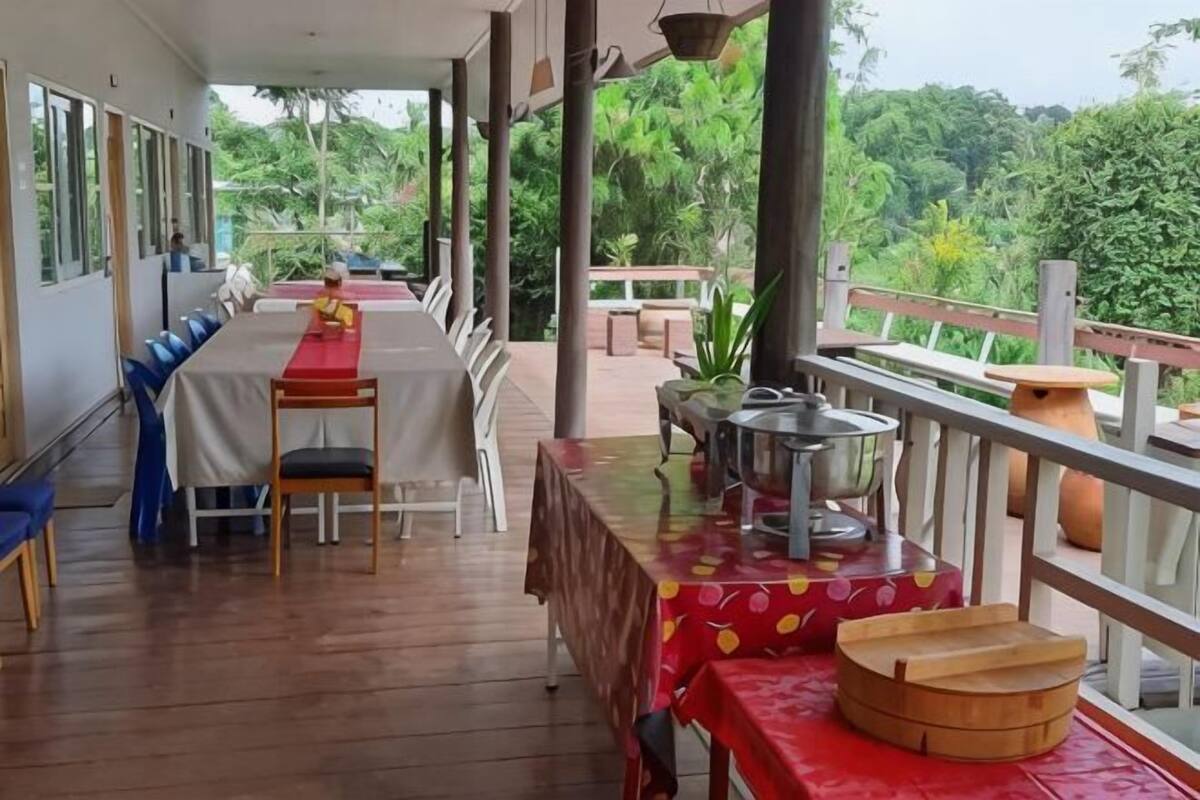Uma área de refeições coberta ao ar livre, com mesas e cadeiras, vista para o verde e uma varanda com plantas em vasos.
