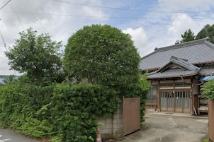 Uma casa japonesa tradicional com telhado de telhas, portão de madeira e uma grande árvore em frente.