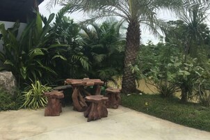 Um pátio de concreto com uma mesa de madeira rústica e banquetas, rodeado por plantas tropicais exuberantes.
