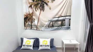 Quarto com uma cama, duas almofadas com a marca 'HEYDAY', uma mesinha de cabeceira pequena e branca, e uma tapeçaria com um carro e palmeiras.