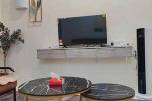 Sala de estar com televisão de tela plana na parede, uma mesa pequena com um objeto vermelho e um aparelho de ar condicionado alto.