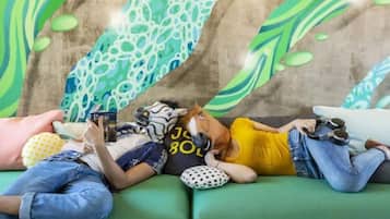 Um sofá verde com um fundo estampado, uma pessoa deitada nele e outra sentada com um laptop.