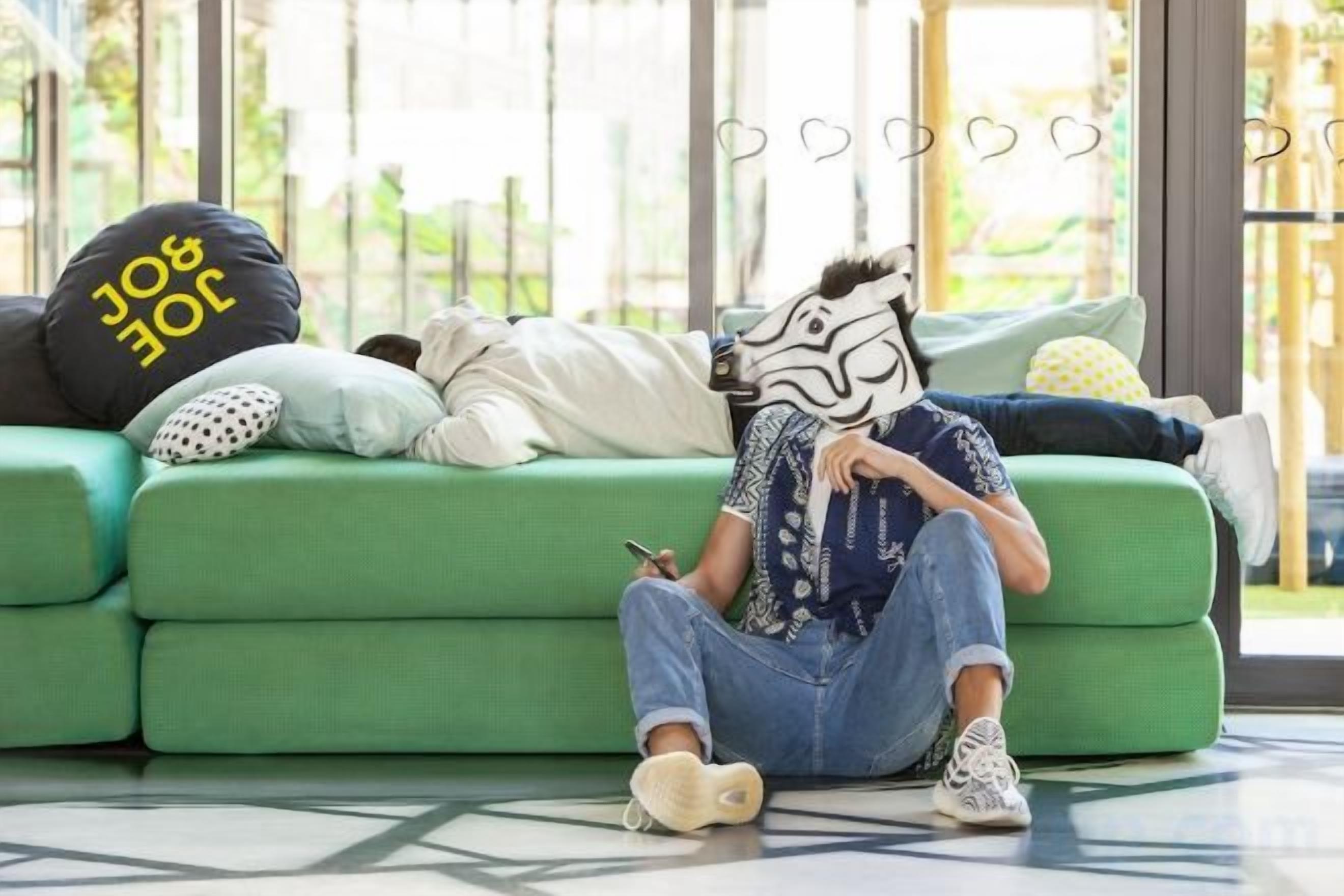 Uma pessoa sentada em um sofá verde com um travesseiro estampado, usando um dispositivo móvel.