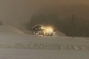 Paisagem coberta de neve com um edifício que tem varanda e janelas iluminadas.