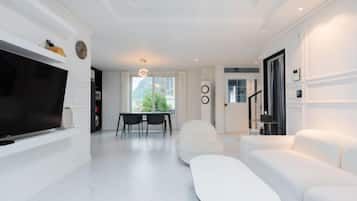 Uma sala de estar moderna com televisão de tela plana, sofás brancos, área de jantar com mesa e cadeiras, e uma janela grande com vista para o verde.