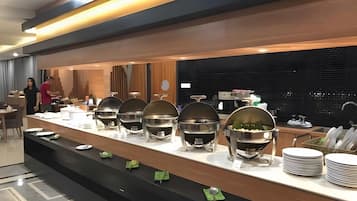 Disposição de buffet com várias estações de serviço, um balcão com pratos empilhados e vista para a cidade através de amplas janelas.