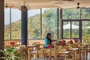 Uma área de refeições com mesas e cadeiras, janelas amplas com vista para colinas e um ventilador de teto.