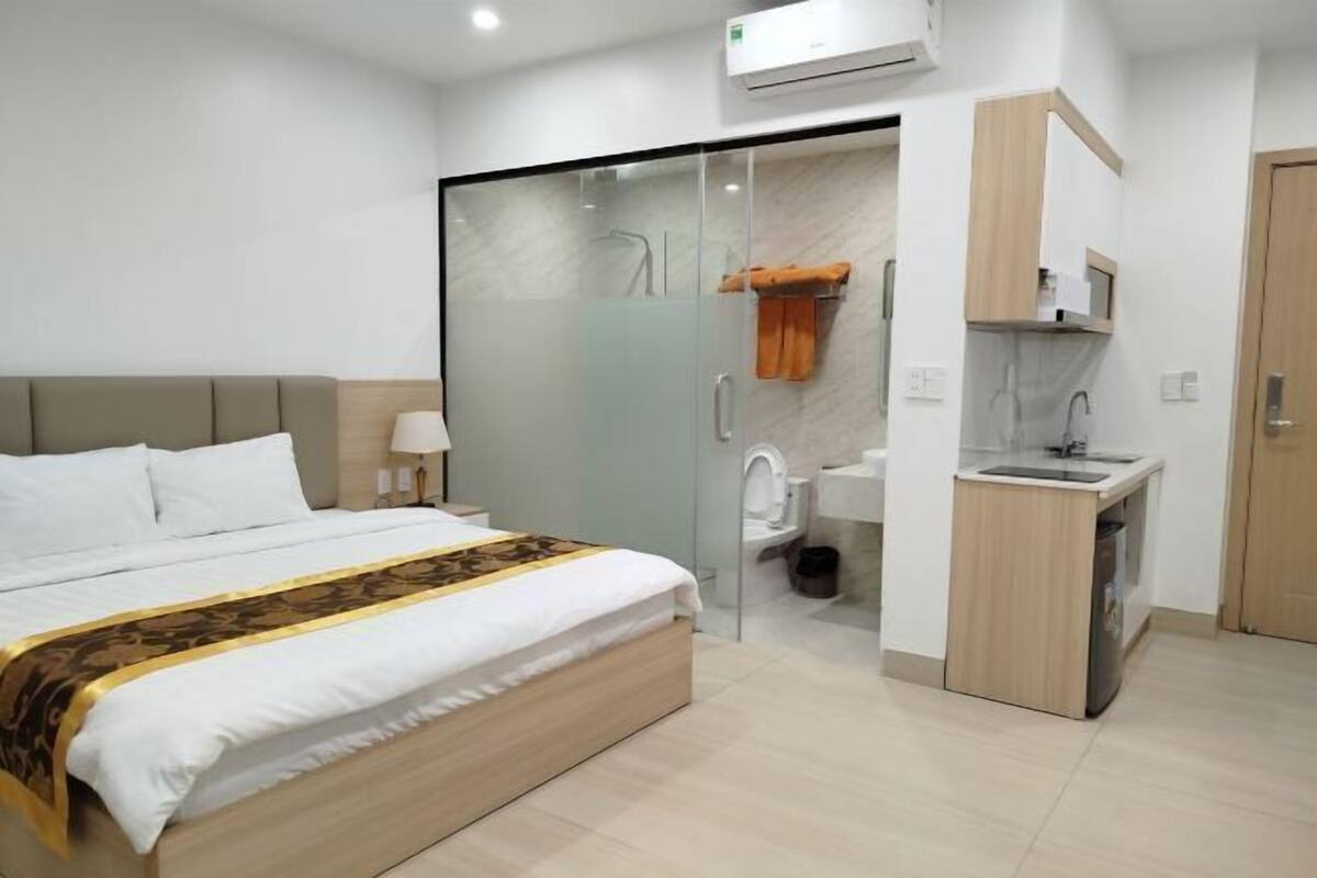 Quarto de hotel moderno com cama, box de vidro para o chuveiro, banheiro pequeno e cozinha compacta.