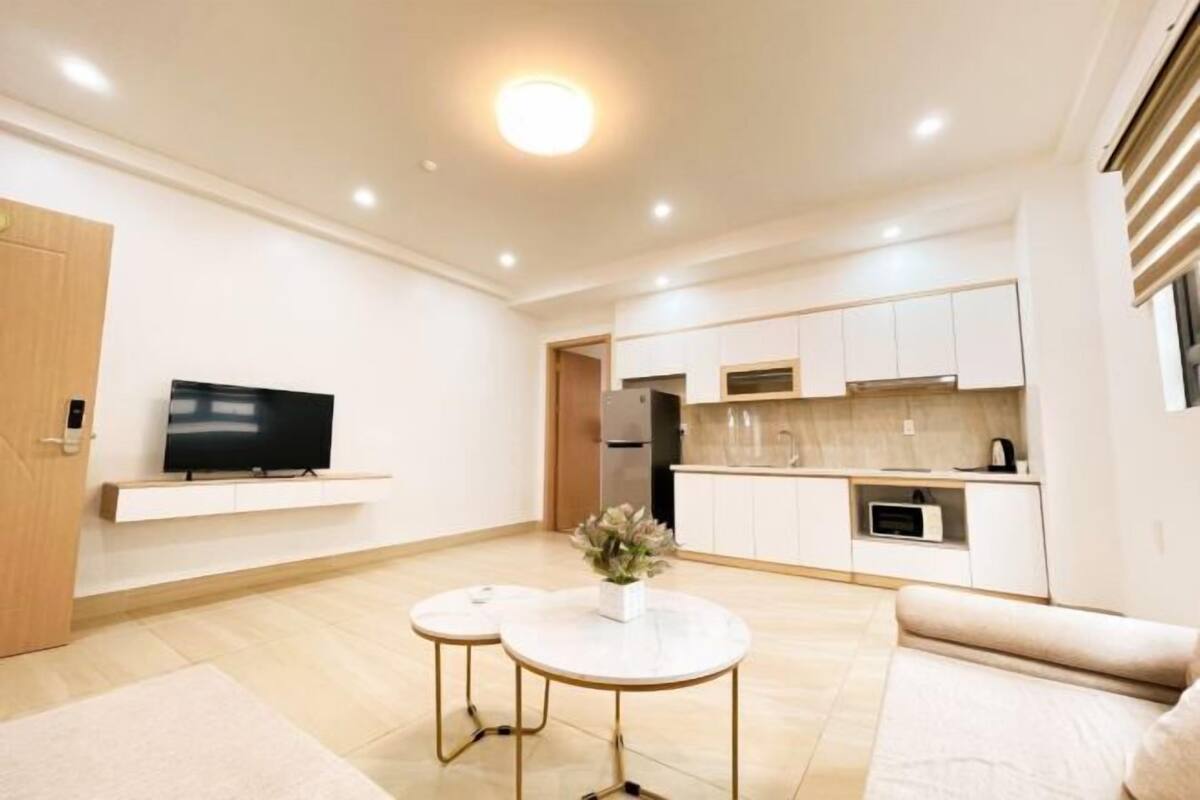 Uma sala de estar moderna com uma televisão de tela plana, duas mesas redondas, um sofá e uma área de cozinha com armários brancos e forno embutido.
