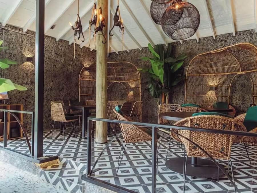 Restaurante com móveis de vime, piso estampado e luminárias pendentes.