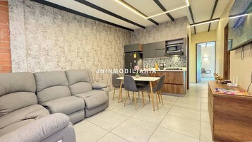 Uma sala de estar moderna com um sofá cinzento, uma área de jantar com mesa e cadeiras, e uma cozinha com micro-ondas e fogão.