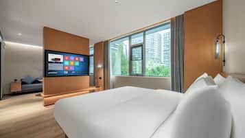 Quarto de hotel moderno com uma cama grande, televisão na parede, janela com vista para edifícios e um pequeno espaço de estar com sofá e mesa lateral.