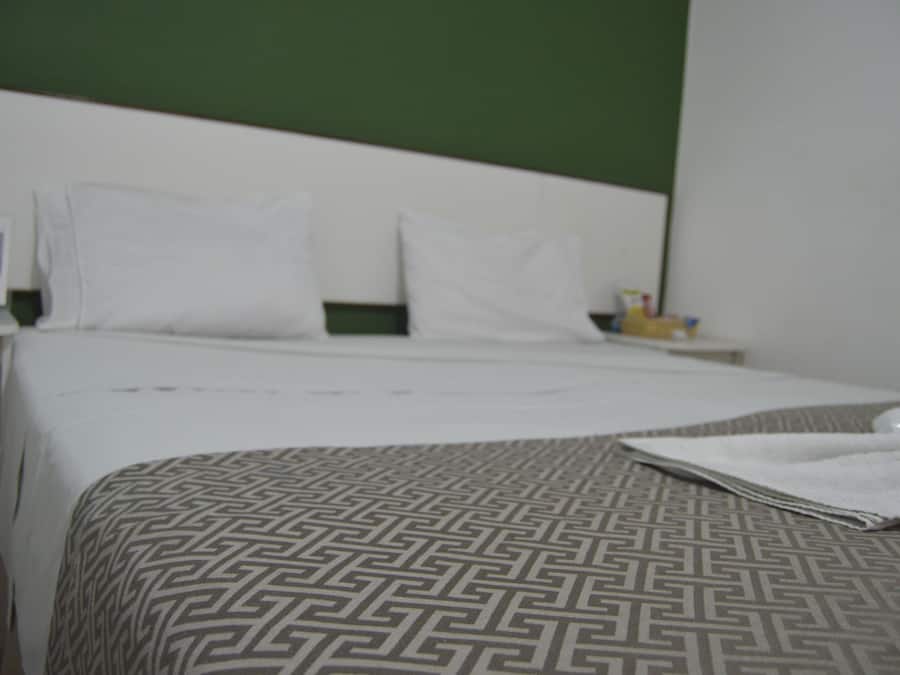Quarto de hotel com uma cama, almofadas brancas, colcha estampada e uma mesa de cabeceira com abajur e uma garrafa.