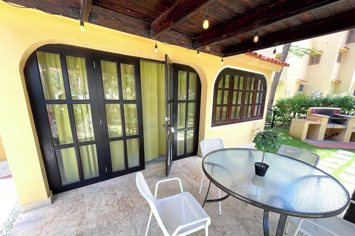 Um pátio externo com uma mesa de vidro redonda e cadeiras brancas, uma porta preta com painéis de vidro e uma parede amarela.