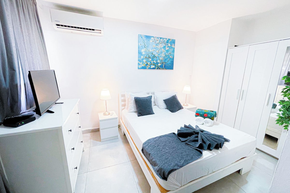 Quarto de hotel moderno com uma cama, mesas de cabeceira, uma escrivaninha com computador e um guarda-roupa.