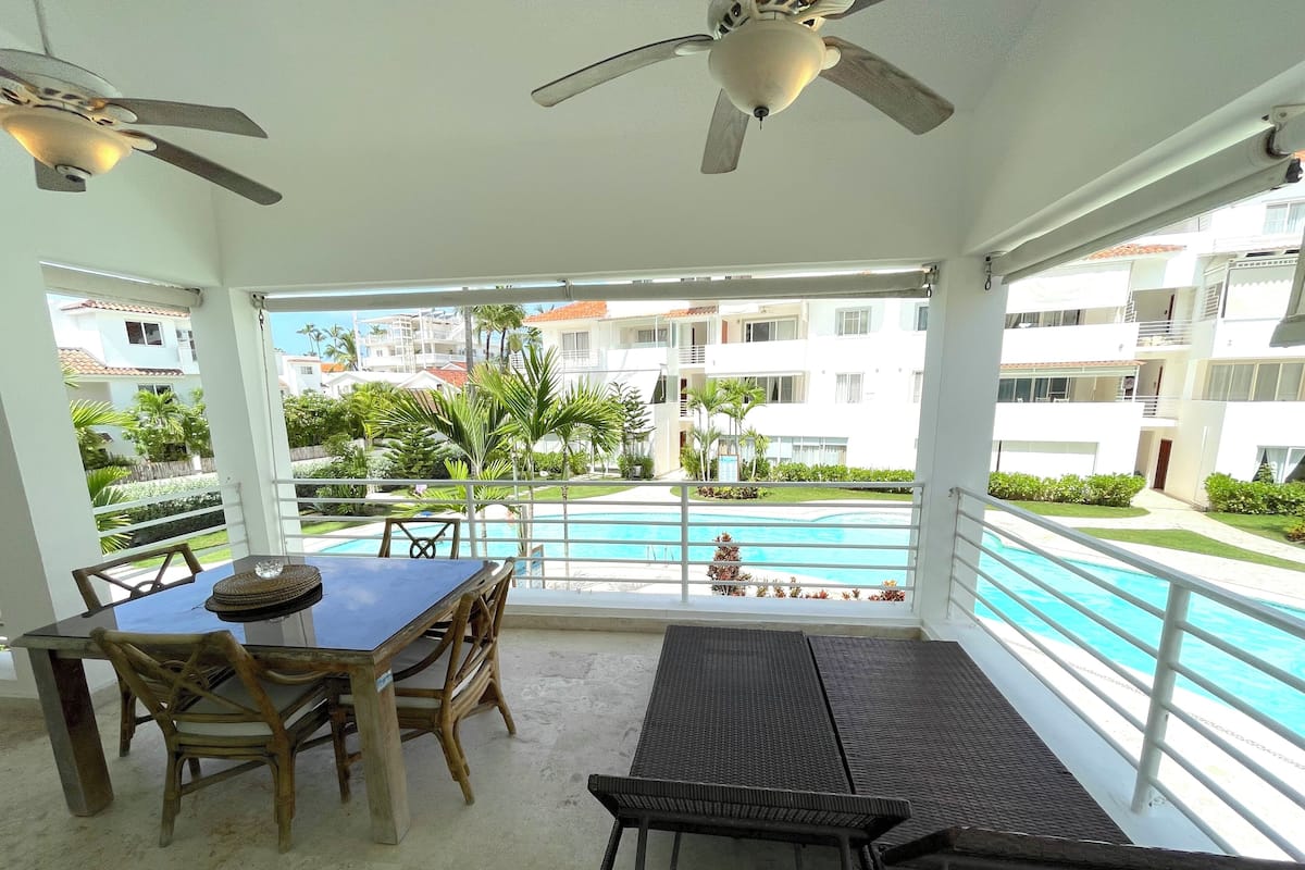 Uma área externa coberta com piscina, mesa e cadeiras, e ventilador de teto.