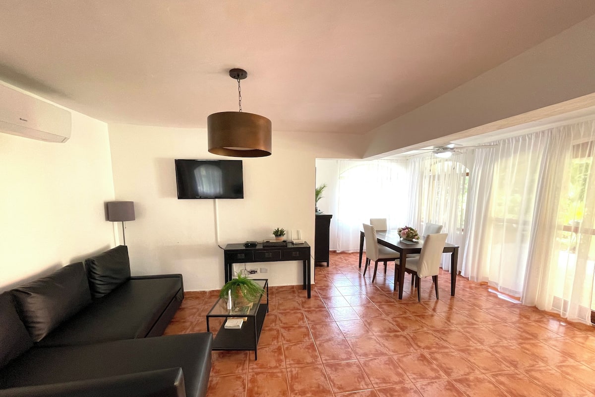 Uma sala de estar com sofá, mesa de centro, televisão e um espaço de jantar com cadeiras e mesa.