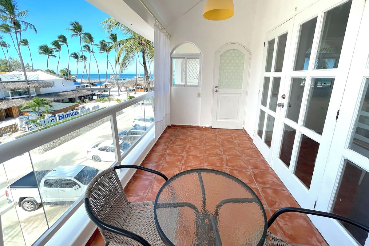Uma varanda com mesa e cadeiras de vidro, com vista para uma praia com palmeiras e uma placa para o 'Blanca Beach Club'.