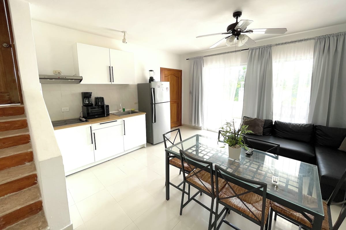 Uma cozinha moderna com armários brancos, geladeira e cafeteira. Há uma área de jantar com mesa e cadeiras de vidro, e um sofá preto com uma planta em cima.