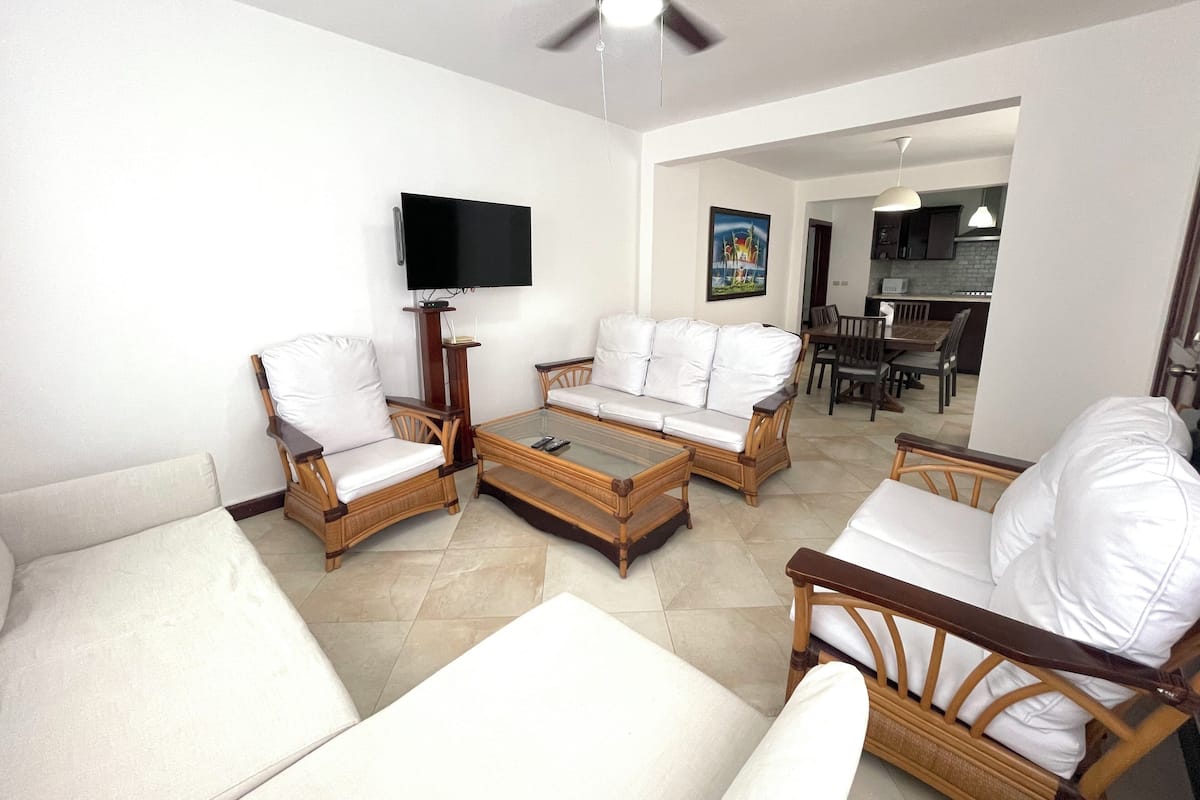Uma sala de estar com ventilador de teto, TV de tela plana na parede e um espaço de jantar com mesa e cadeiras.