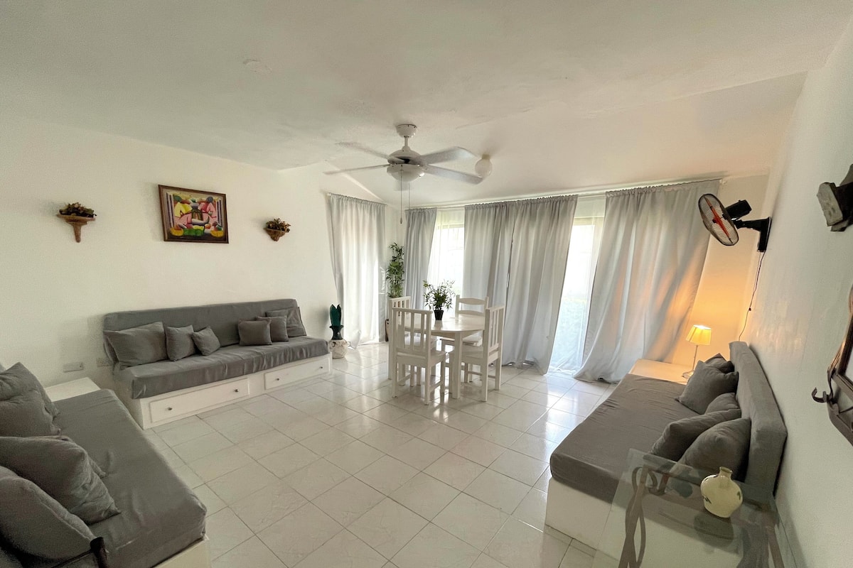 Uma sala de estar ampla com sofá cinza, uma mesa de jantar com cadeiras e um ventilador de teto.