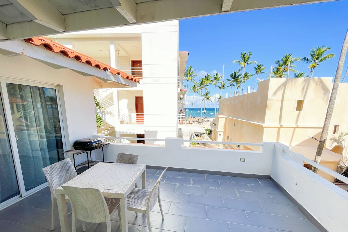 Varanda com mesa e cadeiras, com vista para uma praia e palmeiras.